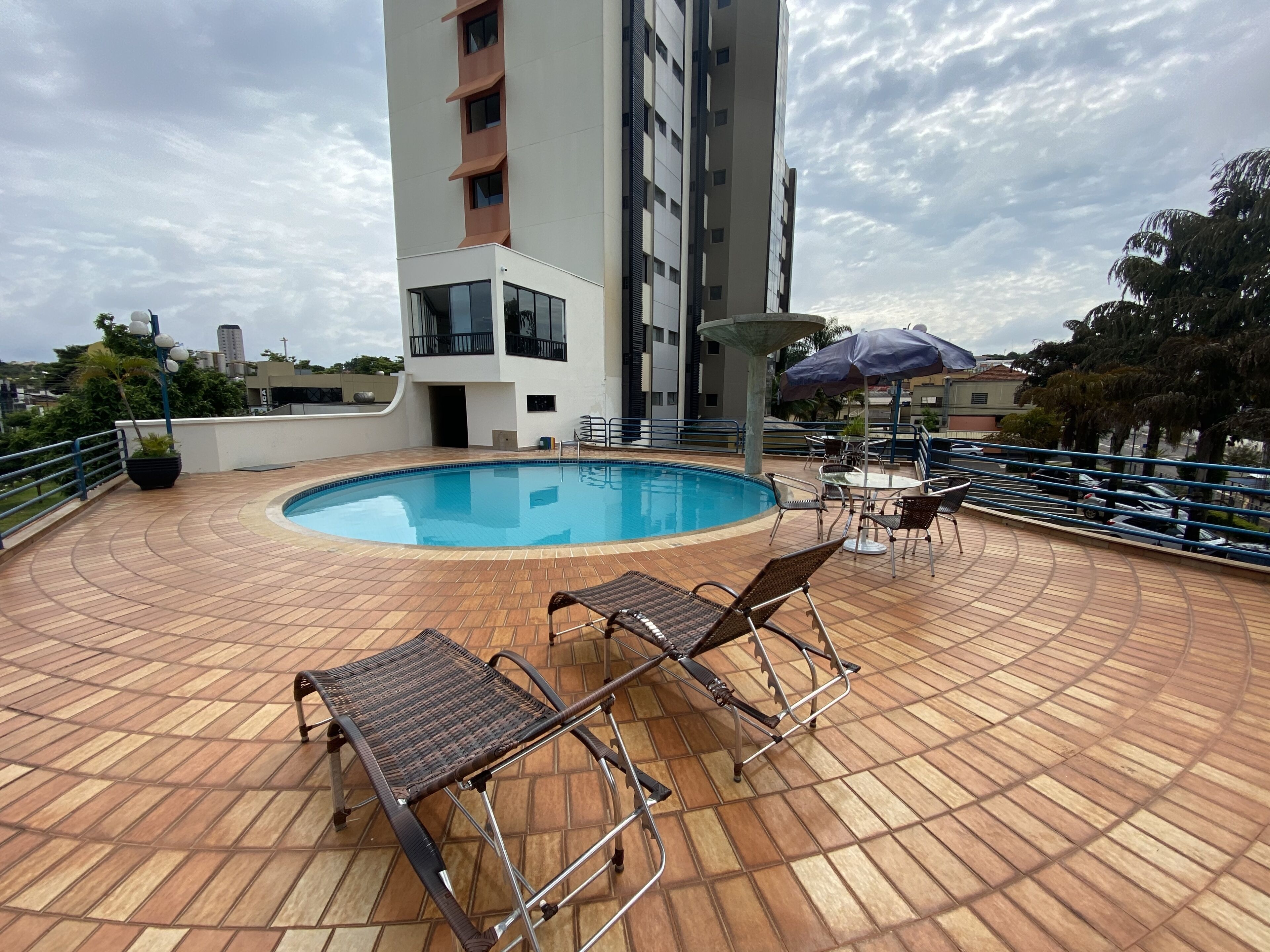 Uma área de piscina no terraço, com espreguiçadeiras e guarda-sóis, cercada por um edifício alto.