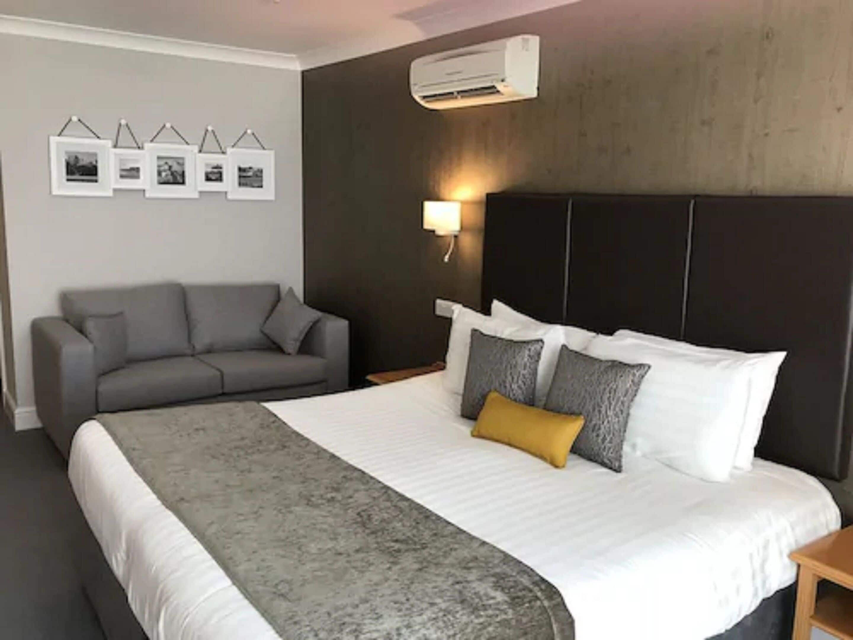 Quarto de hotel com cama, sofá cinzento, ar condicionado fixado na parede e quadros na parede.