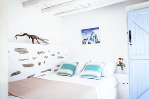 Quarto com parede de pedra, cama com almofadas listradas, pintura de uma paisagem costeira e uma porta azul.