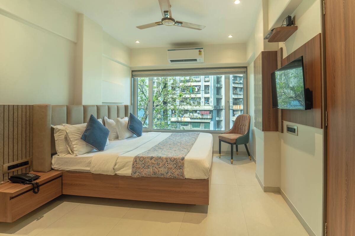 Quarto moderno com cama, mesa de cabeceira, cadeira, TV e janela ampla.