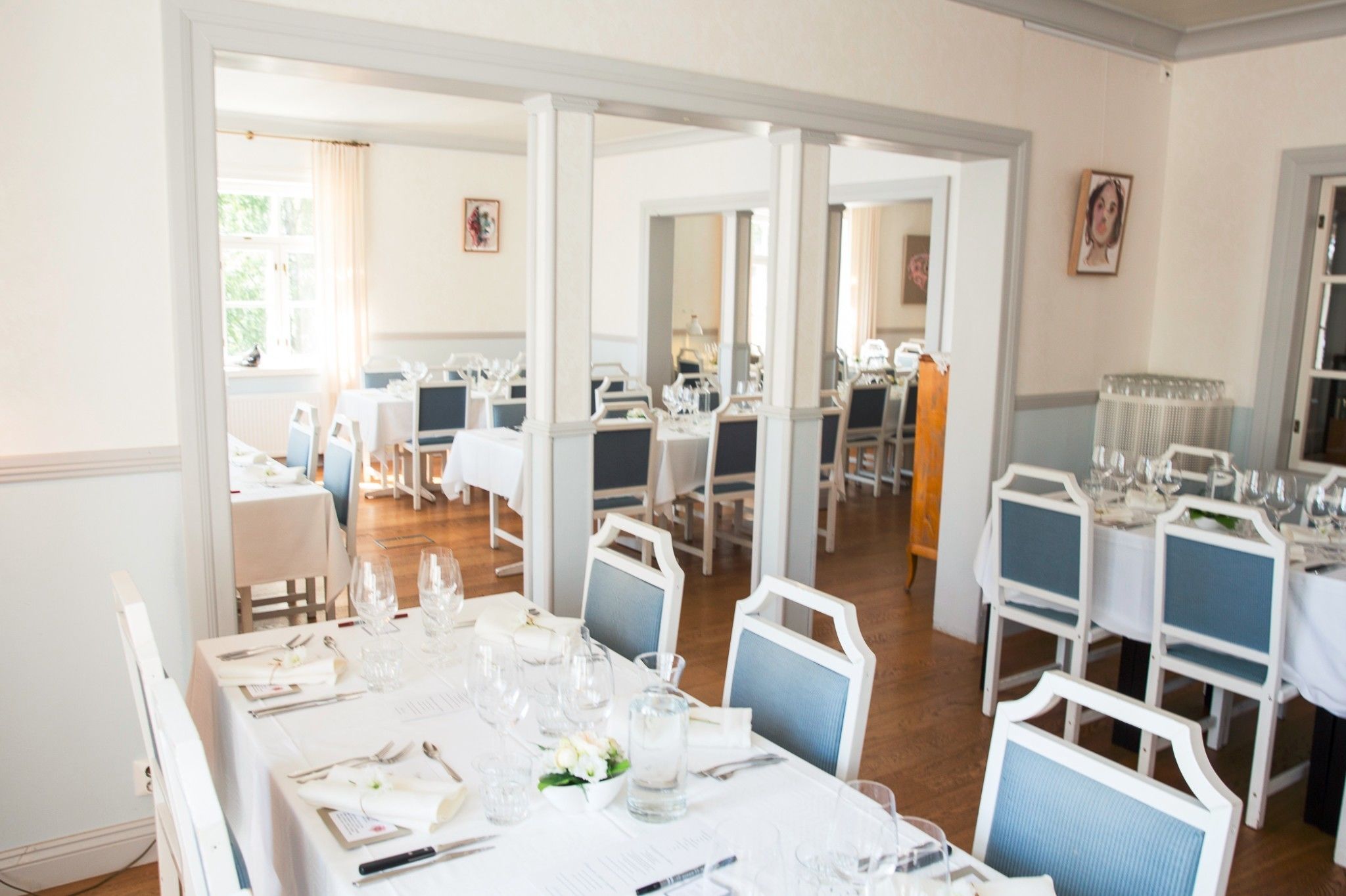 Uma sala de jantar com várias mesas preparadas para o serviço, toalhas brancas e cadeiras azuis.