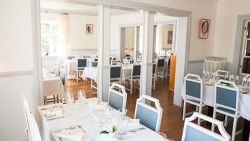 Uma sala de jantar com várias mesas preparadas para o serviço, toalhas brancas e cadeiras azuis.