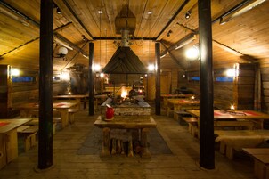 Interior de uma cabana de tronco com paredes e teto de madeira, várias mesas e bancos, uma lareira central e luminárias pendentes.