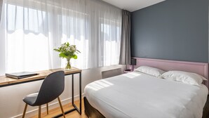 Quarto de hotel com cama, escrivaninha, cadeira e janela com cortinas leves.