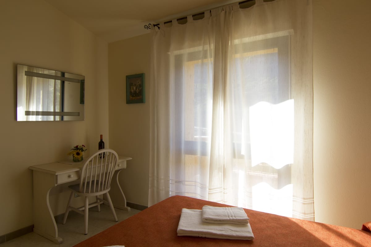Quarto com cama, escrivaninha, cadeira e janela com cortinas leves.