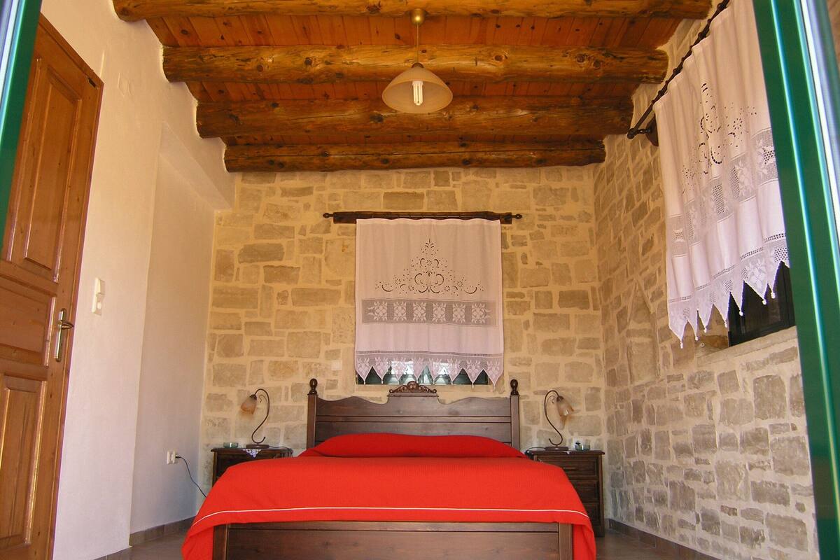 Quarto com parede de pedra, teto de madeira, cama com manta vermelha, mesa de cabeceira, luminária na parede e janela com cortina.
