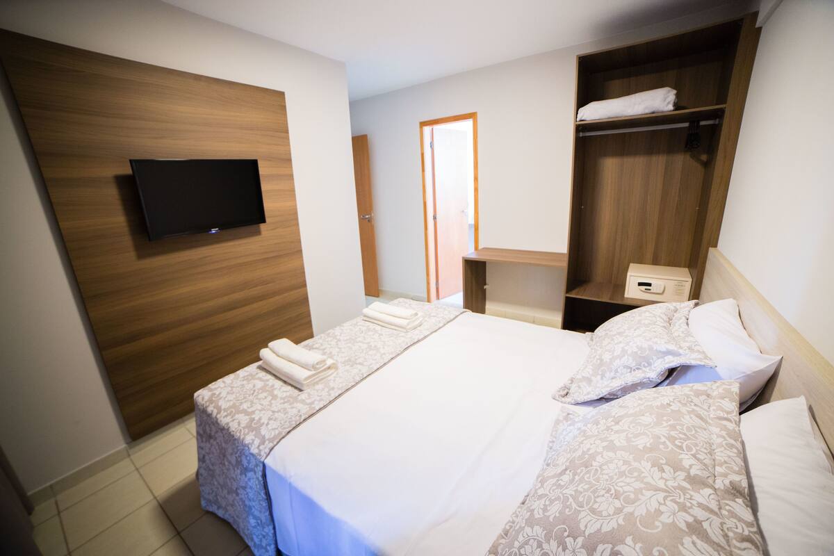 Quarto de hotel com uma cama, televisão fixada na parede, guarda-roupa de madeira e mesa de cabeceira com abajur.