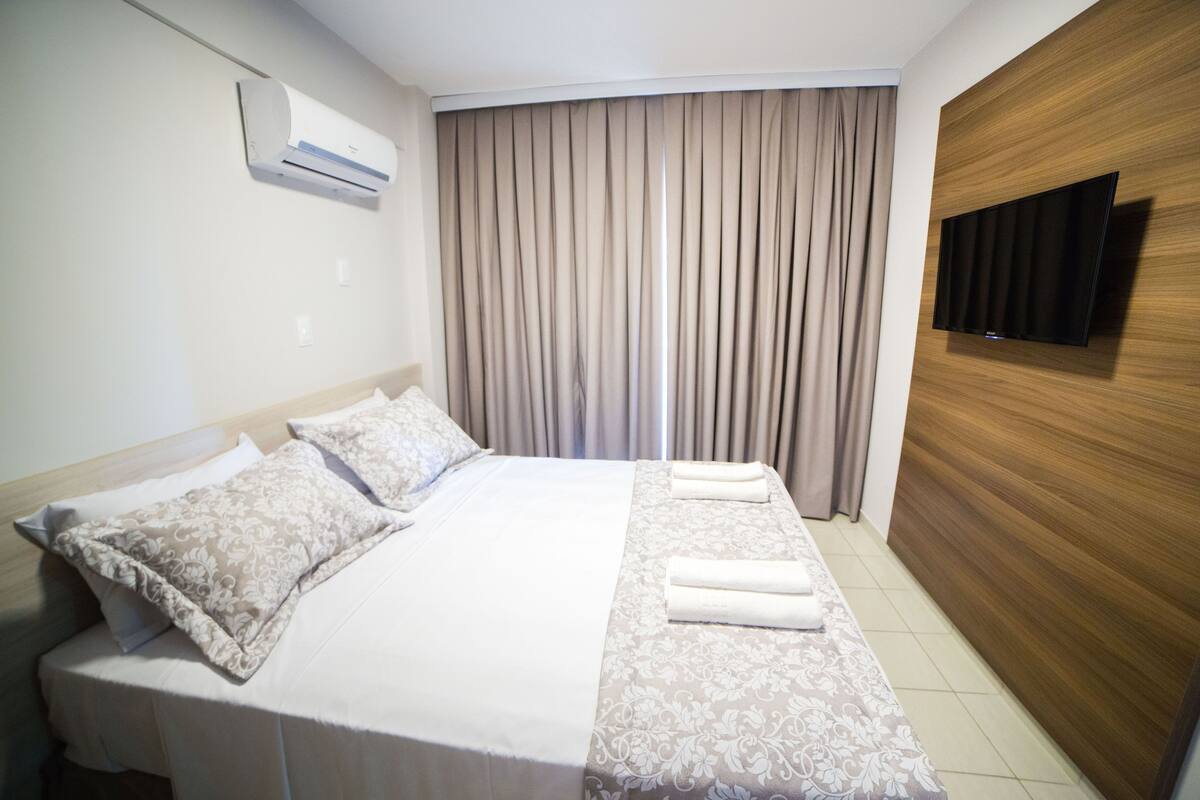 Quarto de hotel com uma cama, uma televisão de tela plana fixada numa parede de madeira e uma janela com cortinas.
