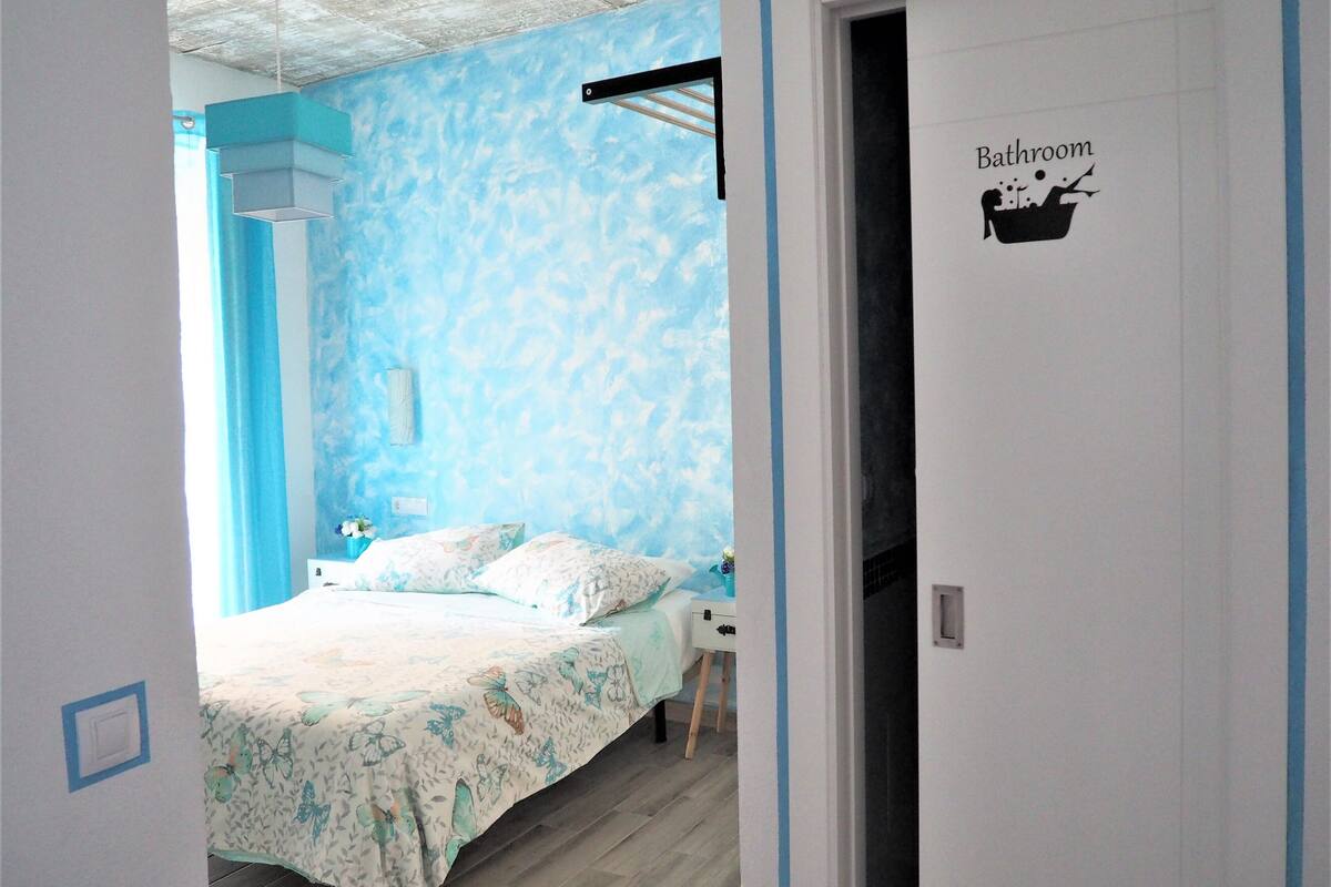 Um quarto com uma cama, uma mesa de cabeceira e uma parede com o símbolo de banheiro.