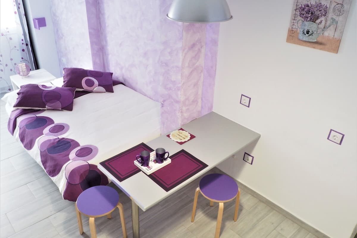 Quarto com cama, escrivaninha, dois banquinhos roxos, luminária e quadro na parede.