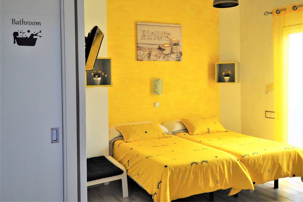 Quarto com duas camas, placa de banheiro, placa com a palavra 'HOME' e uma pequena janela com uma planta.