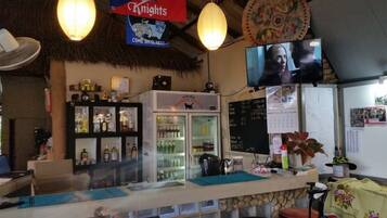 Um bar com teto de palha, balcão com cardápio e uma televisão exibindo um filme.