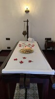 Uma mesa de massagem com toalha branca, bandeja de latão com velas e flores vermelhas.