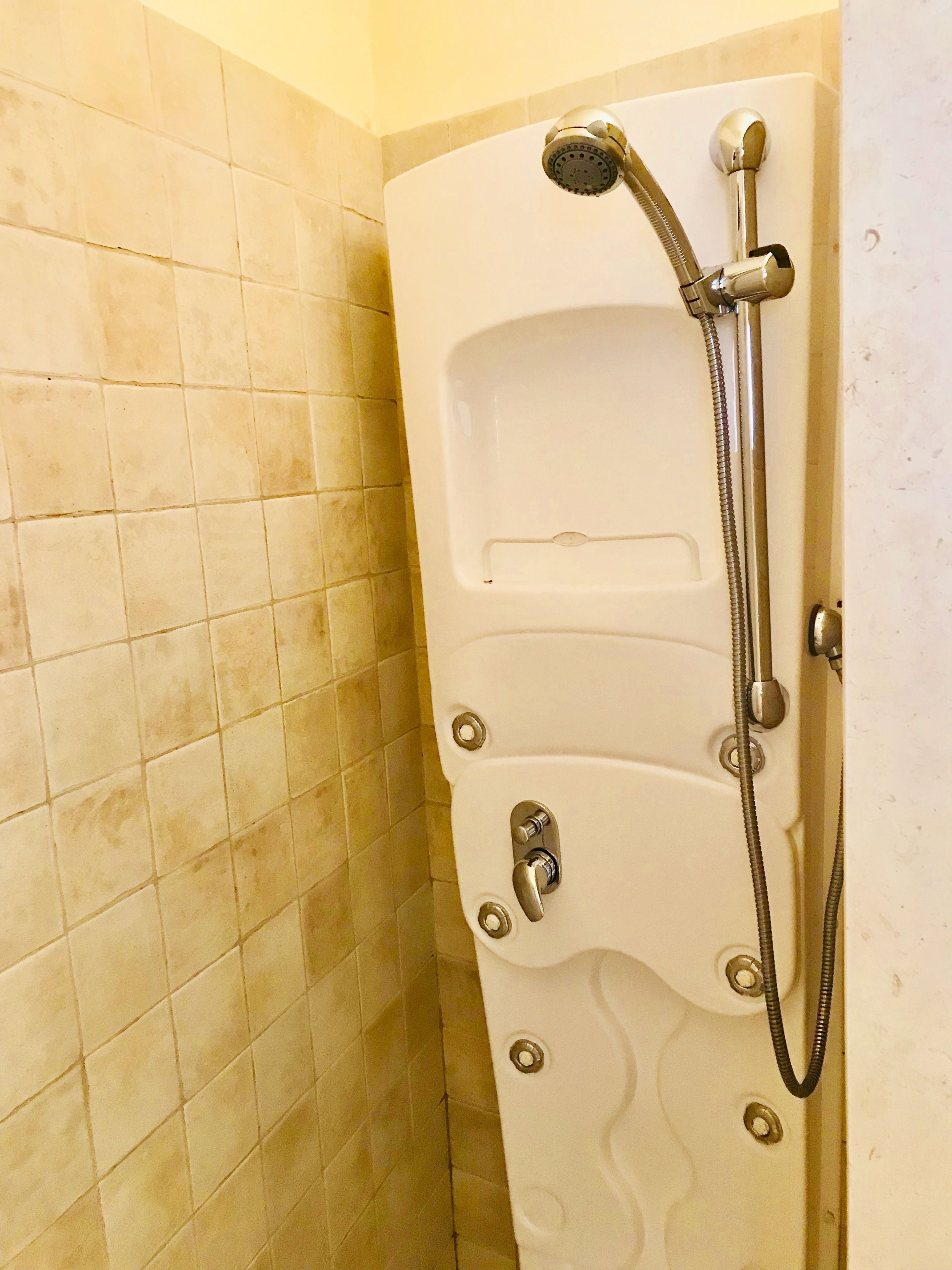Chuveiro com chuveiro manual e fixo, painel de controle e parede revestida de azulejos.