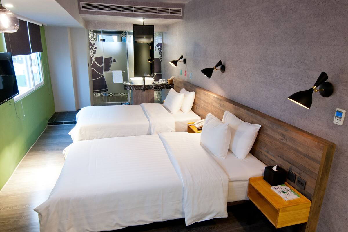 Quarto de hotel com duas camas, cabeceira de madeira, luminárias de apoio, espelho, pia e televisão.