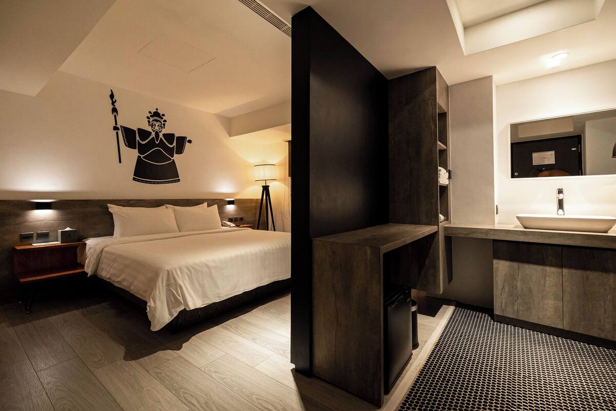 Quarto de hotel moderno com uma cama grande, uma mesa de cabeceira de madeira, uma TV fixada na parede e um banheiro com um espelho grande e pia.