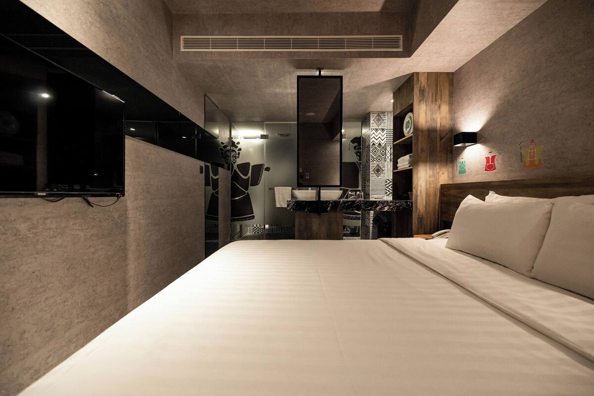 Quarto de hotel moderno com uma cama grande, televisão integrada, toucador com espelho e zona de chuveiro.