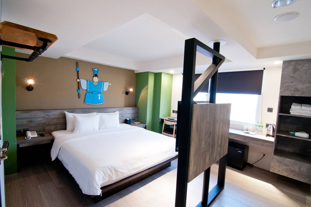 Quarto de hotel moderno com uma cama grande, uma escrivaninha com computador, uma kitchenette pequena e uma parede de destaque verde.