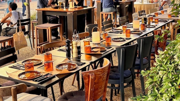 Um café ao ar livre com mesas, cadeiras e um bar, com vista para a rua.