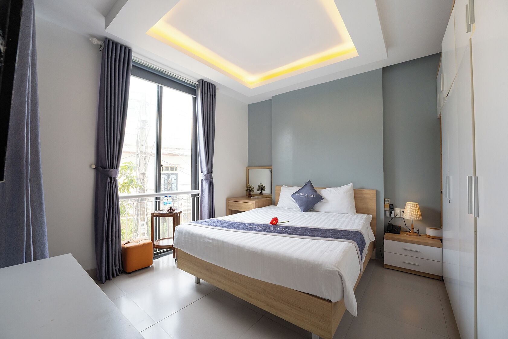 Quarto de hotel moderno com uma cama grande, mesas de cabeceira, luminária e vista da cidade através de amplas janelas.