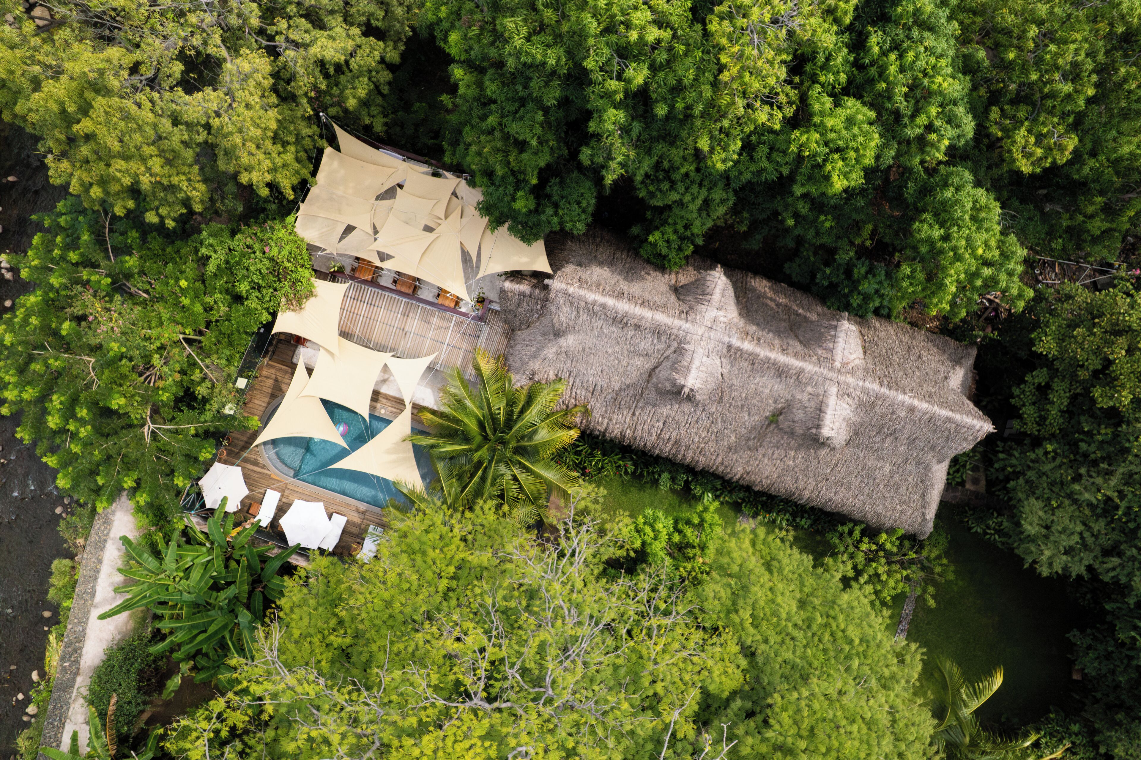 Vista aérea de um resort com estrutura de telhado de palha, área de piscina com guarda-sóis e vegetação exuberante ao redor da propriedade.