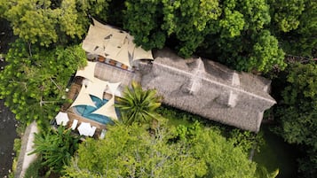 Vista aérea de um resort com estrutura de telhado de palha, área de piscina com guarda-sóis e vegetação exuberante ao redor da propriedade.