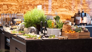 Disposição de buffet com diversos alimentos, um arranjo central de planta verde grande e um painel de fundo com um mural detalhado.