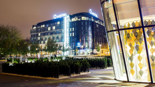 Um hotel moderno com uma placa grande 'QHOTEL', fachada de vidro e entrada bem iluminada.