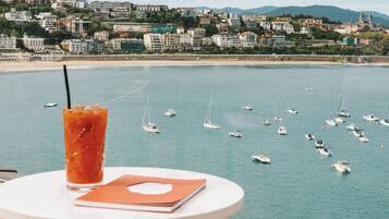 Mesa com bebida e menu, com vista para um porto com barcos e uma cidade costeira ao fundo.