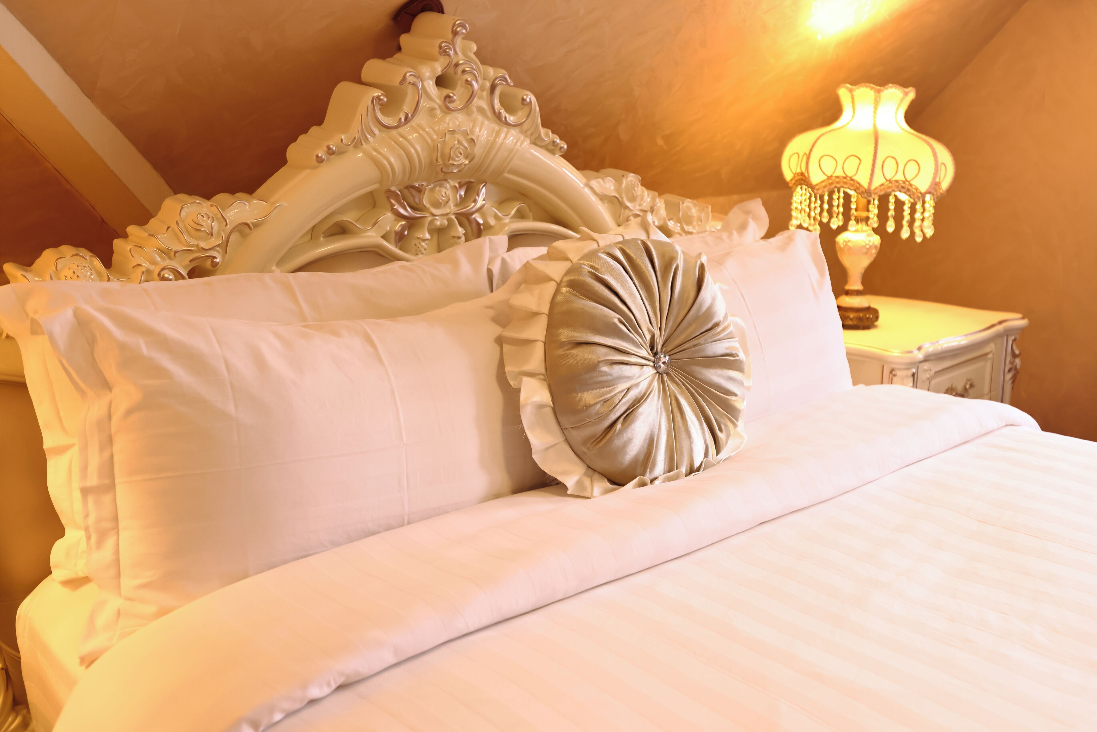 Cama bem arrumada com lençóis brancos e almofadas decorativas, uma cabeceira clássica e um abajur.