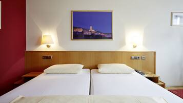 Quarto de hotel com duas camas, uma cabeceira de madeira, luminárias laterais, um quadro com vista urbana e uma parede em destaque na cor vermelha.