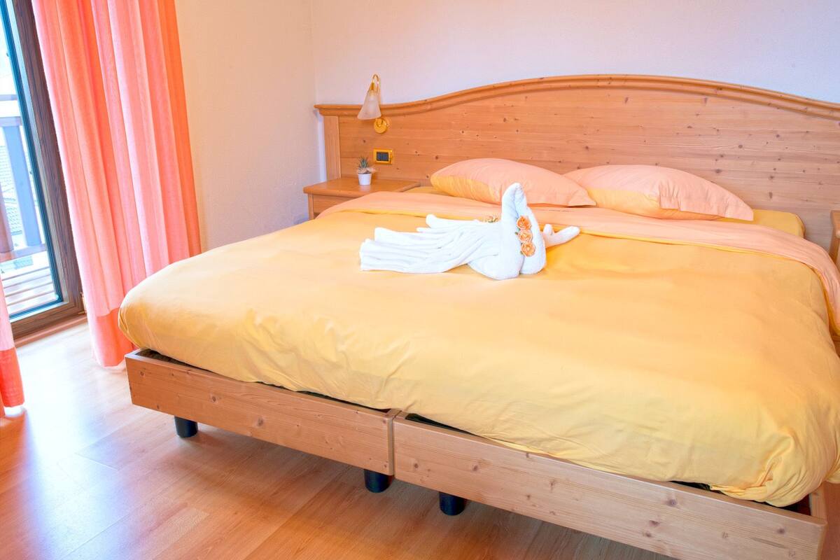 Uma cama de madeira com colcha amarela, um criado-mudo de madeira com uma lâmpada e uma janela com cortinas.