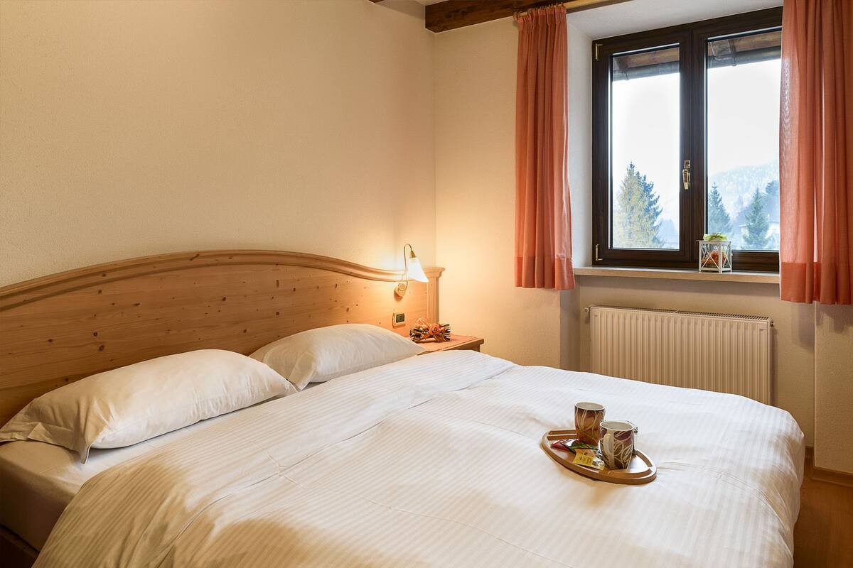 Quarto com cabeceira de madeira, mesa de cabeceira com abajur, janela com cortinas e bandeja com caneca e xícara sobre a cama.