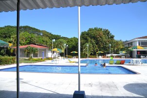 Piscina com espreguiçadeiras e guarda-sóis, cercada por vegetação e um edifício ao fundo.