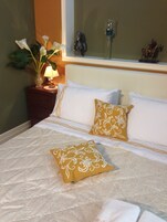 Cama bem arrumada com almofadas brancas e estampadas, mesa de cabeceira com abajur e um vaso com flores.
