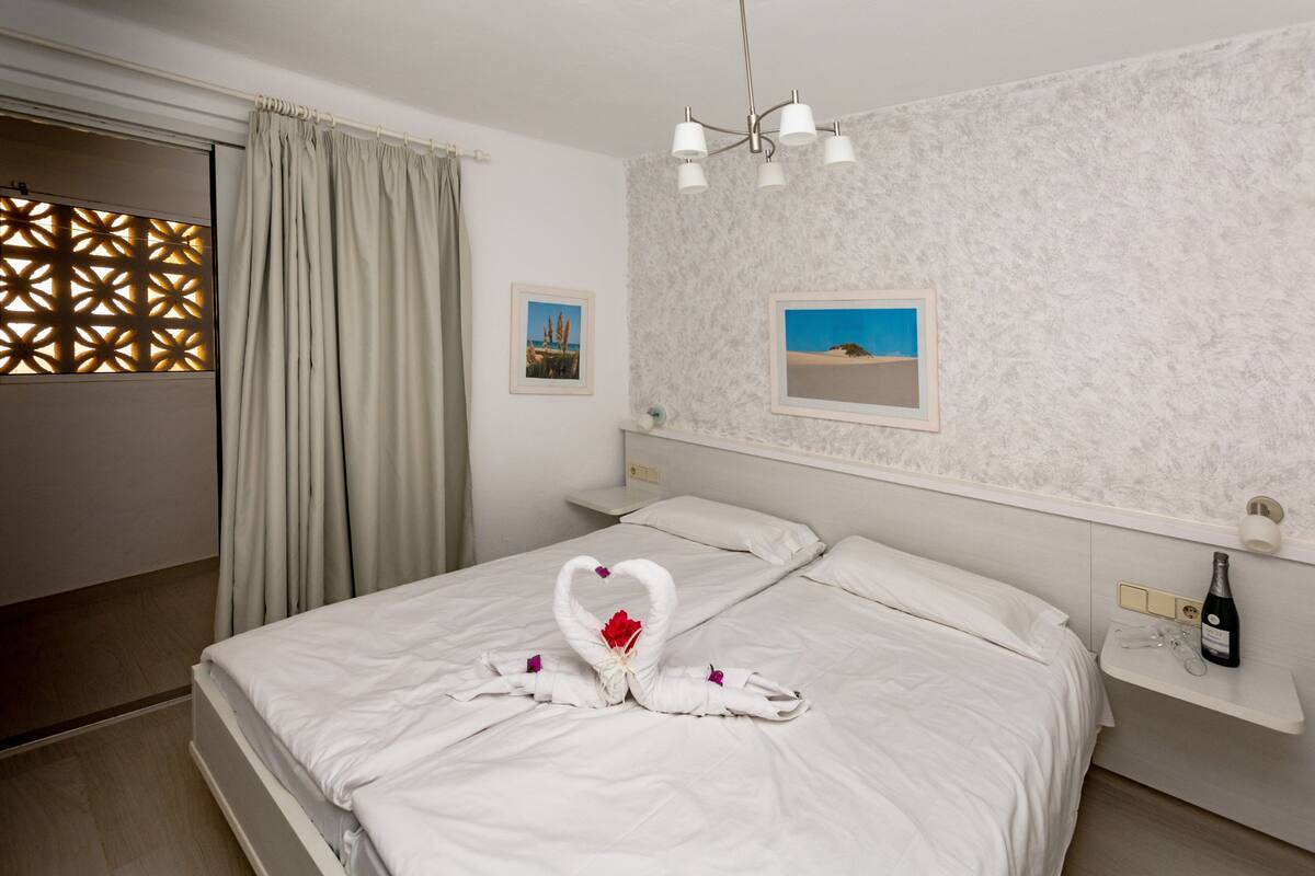 Quarto de hotel com cama, mesa de cabeceira, prateleira na parede e janela com cortinas.