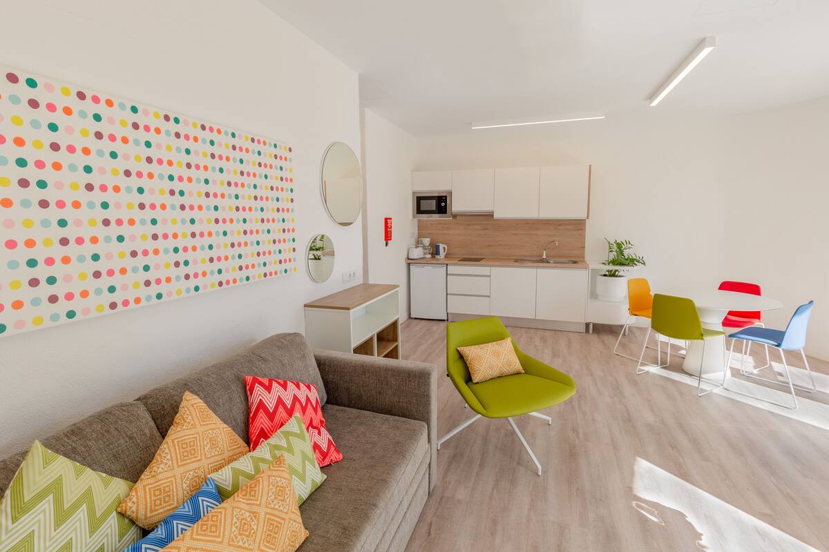 Uma sala de estar moderna com um sofá, almofadas coloridas, uma poltrona verde e uma área de cozinha com armários brancos e um micro-ondas.