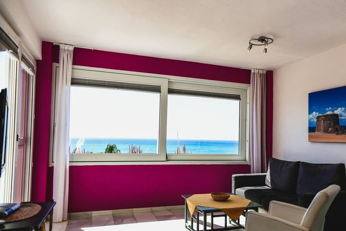 Quarto com uma janela grande que oferece vista para o mar, uma mesa com uma tigela, um sofá e uma luminária no teto.