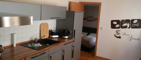 Uma cozinha compacta com armários cinza, bancada de madeira, pia, cafeteira e geladeira. Uma cama é visível no quarto adjacente.