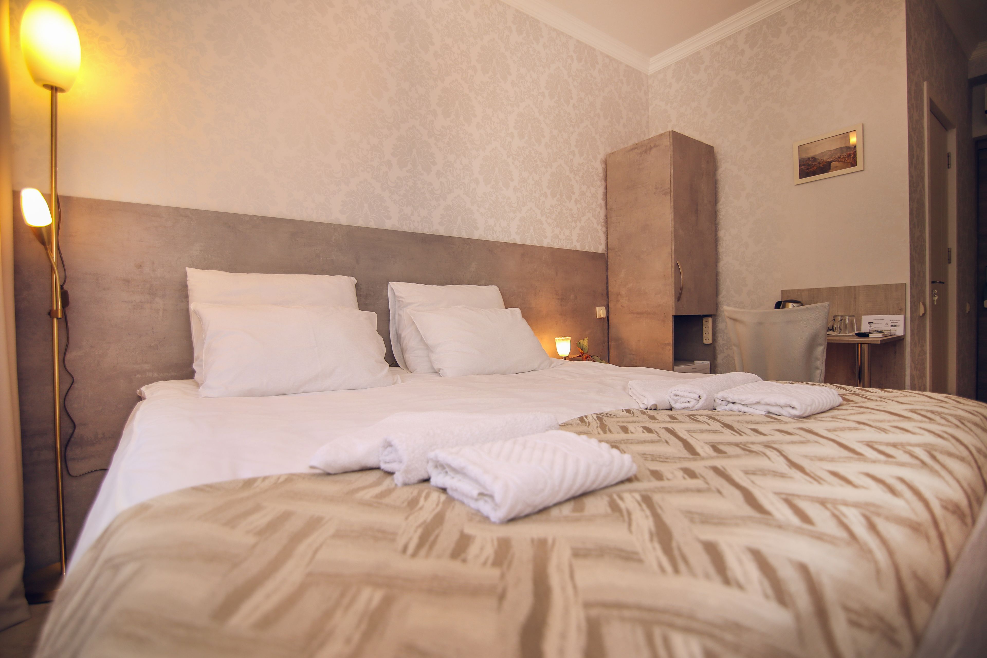 Quarto de hotel com uma cama grande, uma mesinha de cabeceira, uma luminária e um guarda-roupa de madeira.