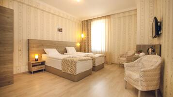 Quarto de hotel com duas camas, cabeceira de madeira, mesa de cabeceira com luminária, uma cadeira, pequena mesa com espelho e janela com cortinas.