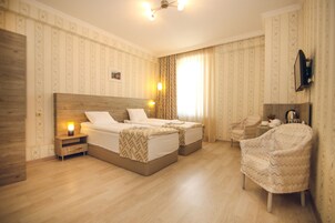 Quarto de hotel com duas camas, cabeceira de madeira, mesa de cabeceira com luminária, uma cadeira, pequena mesa com espelho e janela com cortinas.