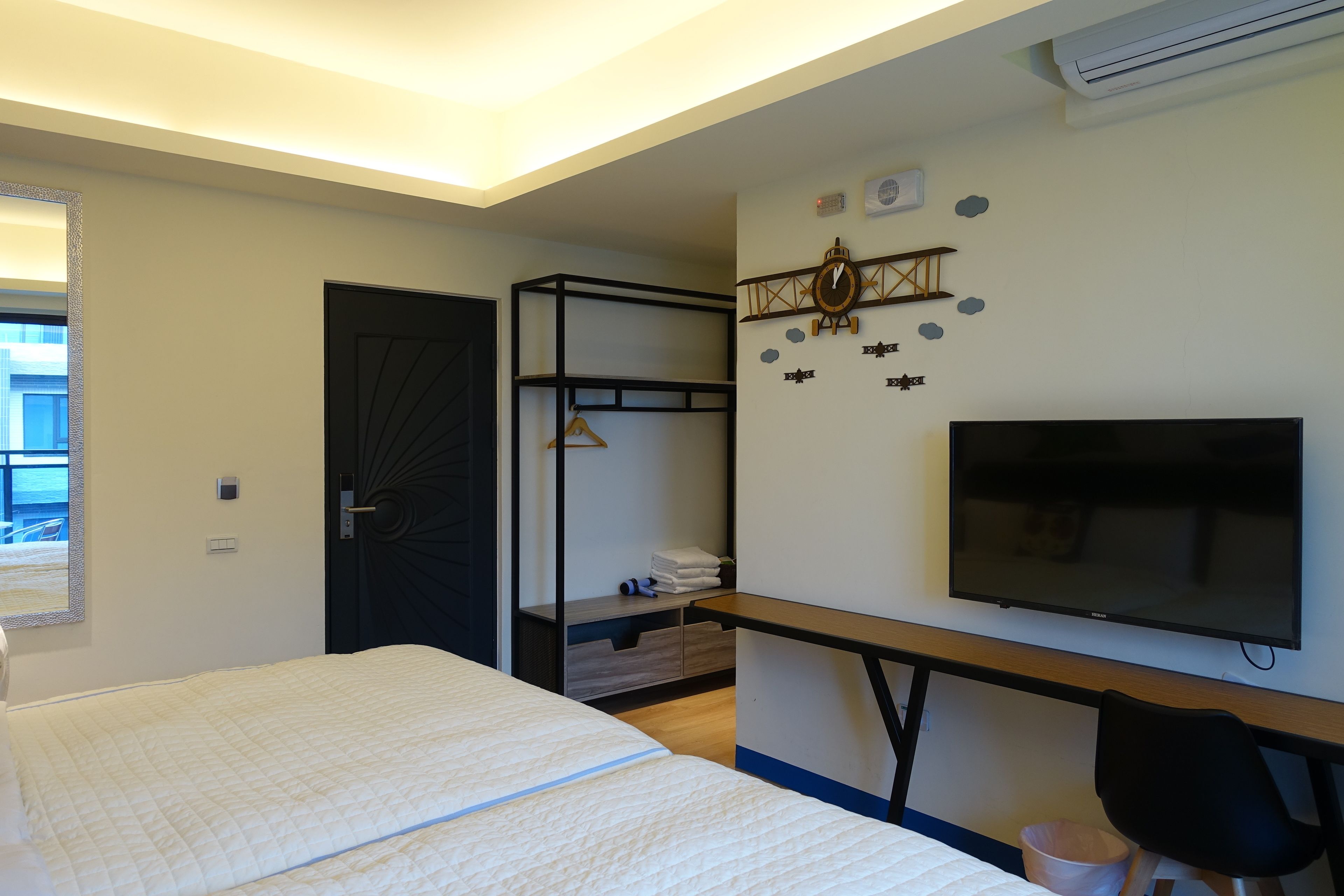 Quarto de hotel com cama, escrivaninha com televisão, beliche e relógio de parede.