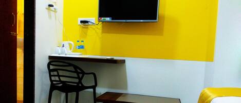 Quarto de hotel pequeno com escrivaninha, cadeira e televisão fixada na parede.