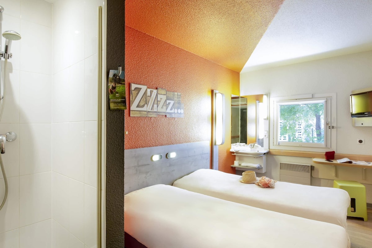 Quarto de hotel com duas camas, chuveiro, pia e janela com vista para árvores.