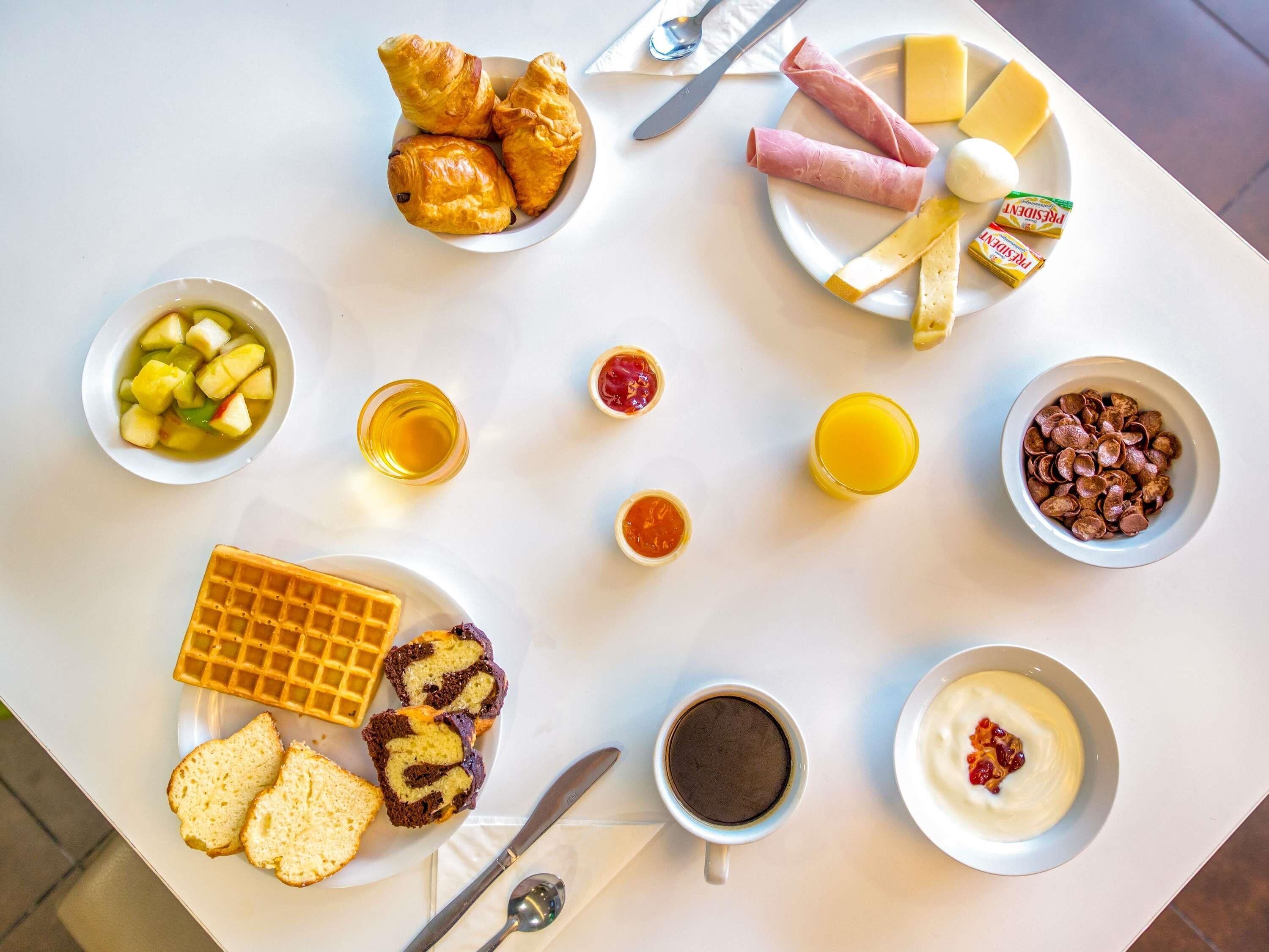 Mesa de café da manhã com croissants, presunto, queijo, frutas, waffles, chocolate e bebidas.
