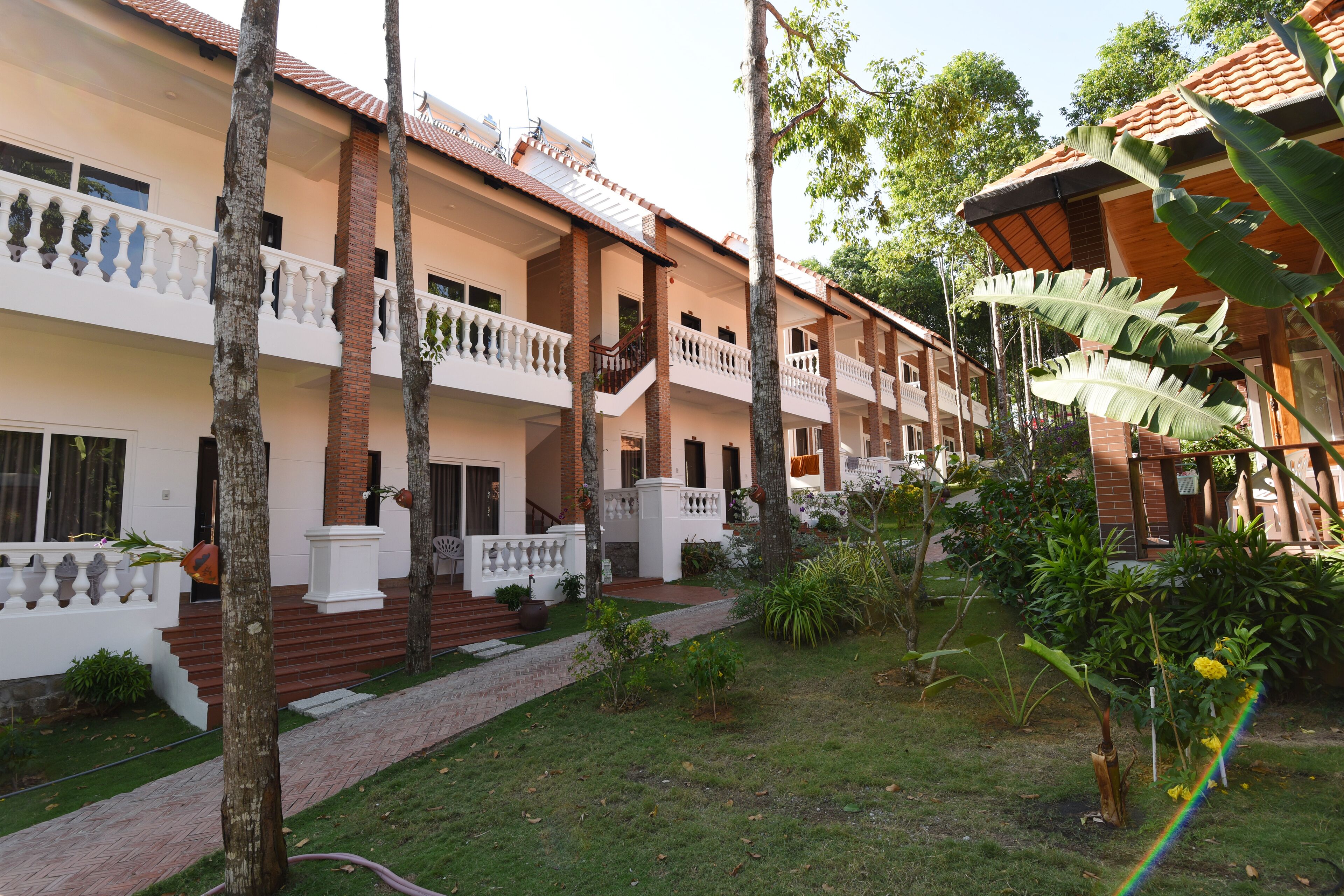 Hotel de vários andares com varandas, fachada de tijolos e área de jardim com plantas tropicais.