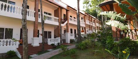 Hotel de vários andares com varandas, fachada de tijolos e área de jardim com plantas tropicais.