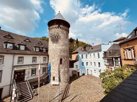 Uma torre de pedra histórica com telhado cônico, rodeada por edifícios europeus tradicionais com telhados inclinados e estrutura de madeira.