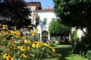 Um edifício branco com persianas verdes, rodeado por vegetação exuberante e flores amarelas.